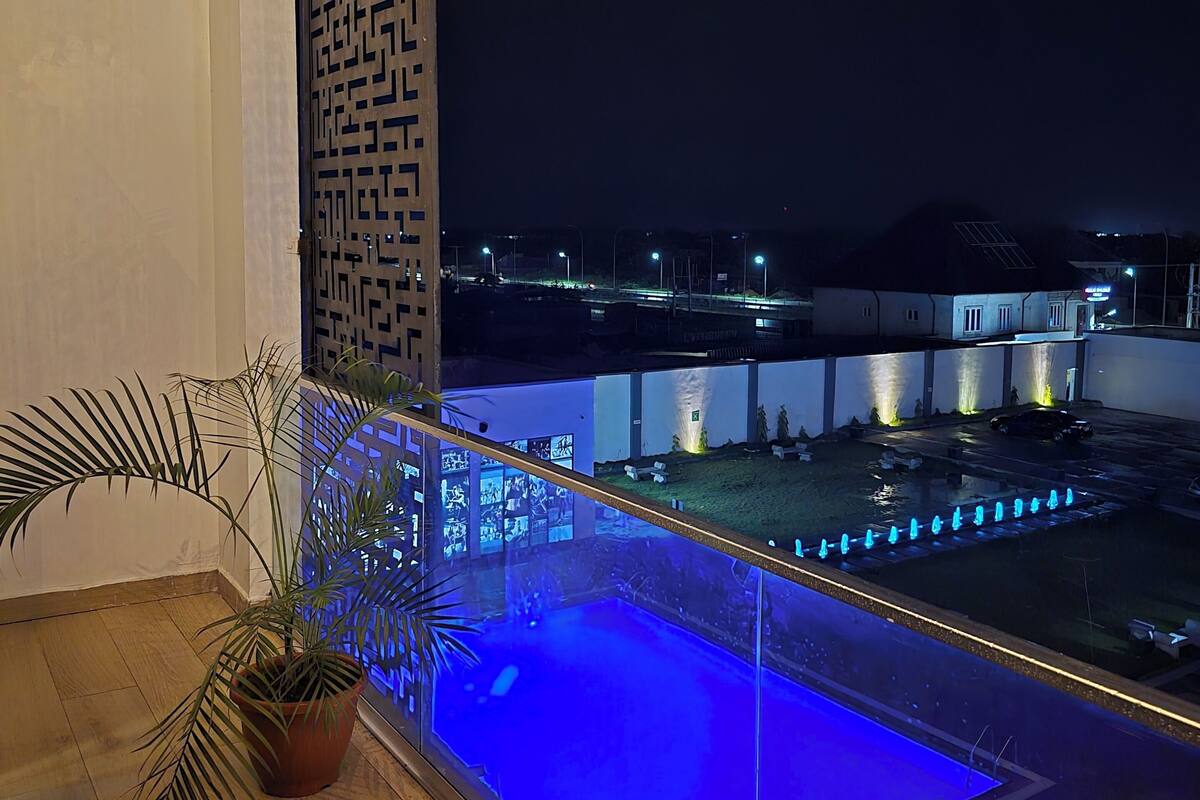 Varanda com piscina, plantas em vasos e vista para área iluminada com fontes e um carro.
