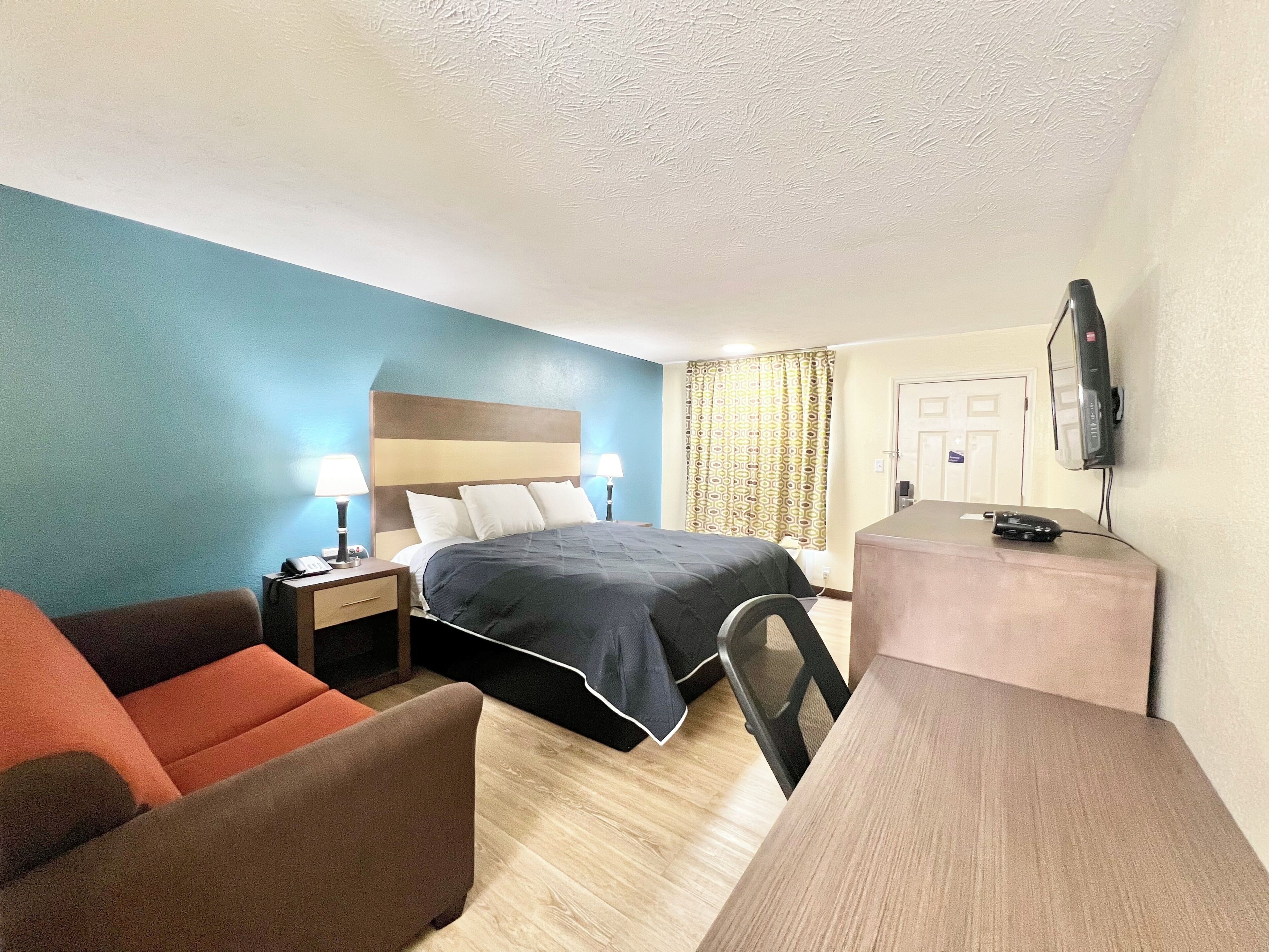 Quarto de hotel com cama, escrivaninha, cadeira, mesa de cabeceira e televisão.