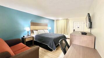 Quarto de hotel com cama, escrivaninha, cadeira, mesa de cabeceira e televisão.