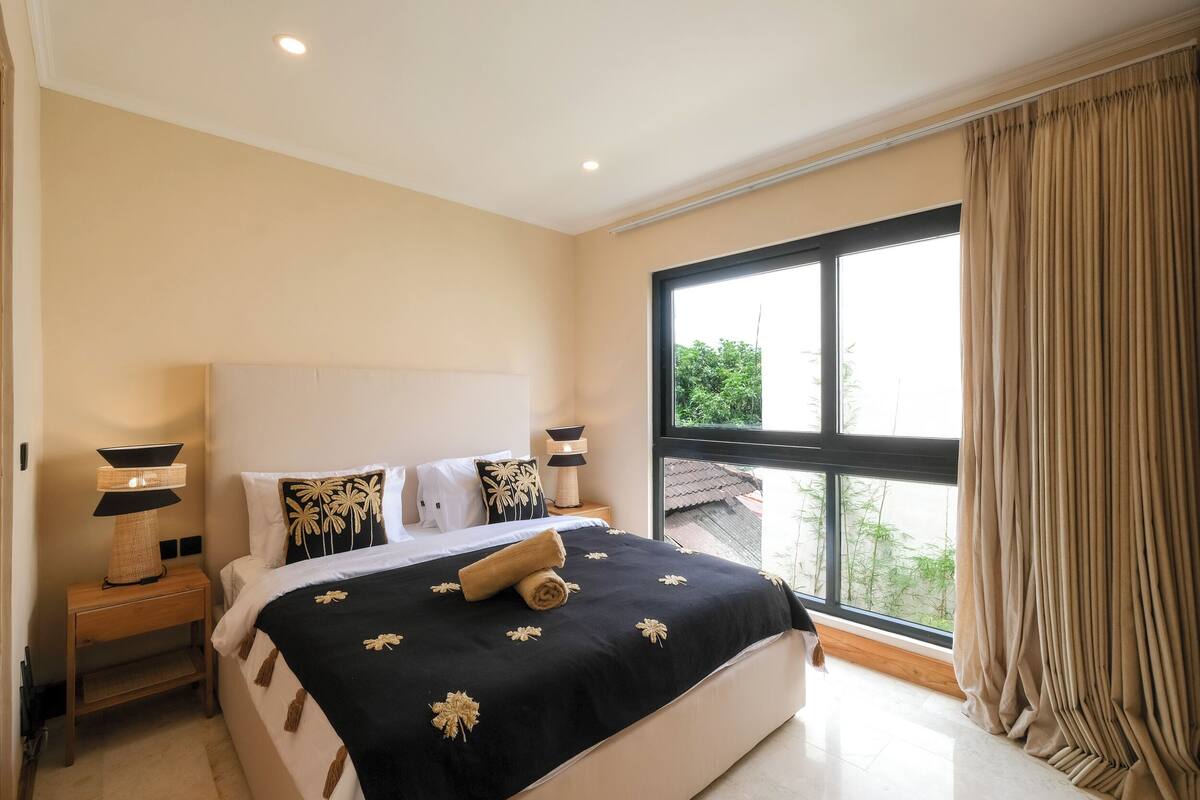 Quarto com cama grande, mesa de cabeceira, luminária e janela com cortinas.