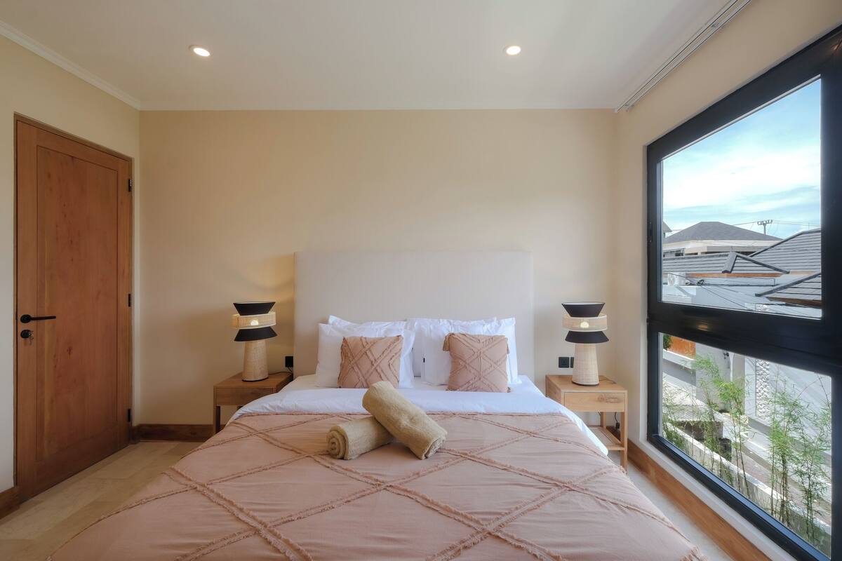 Quarto com uma cama grande, duas mesas de cabeceira com luminárias, uma porta de madeira e uma janela ampla com vista para edifícios.