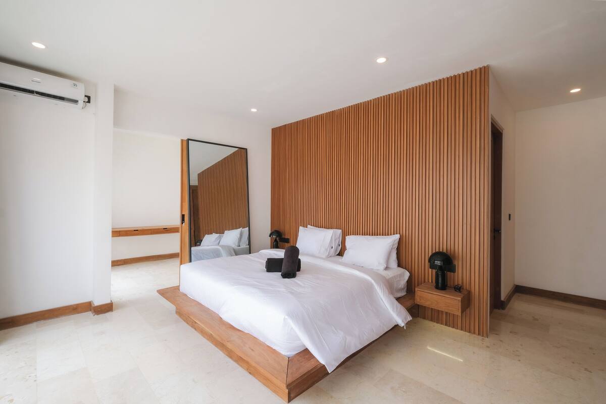 Quarto moderno com cama grande, painel de madeira e pequena mesa de cabeceira com abajur.