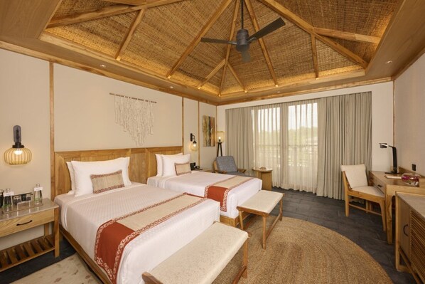 Quarto de hotel com duas camas, uma escrivaninha, uma cadeira, um ventilador de teto e uma janela grande com cortinas.