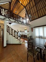 Uma ampla área de jantar com uma mesa grande e cadeiras, uma cozinha ao fundo e uma escada com grades ornamentadas.