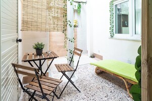 Uma pequena área de pátio com mesa e cadeiras de madeira, uma cama pequena com colchão verde e uma janela com vista para o exterior.