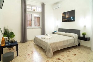 Quarto com cama, mesa de cabeceira, janela com cortinas, ar condicionado fixado na parede e um quadro.