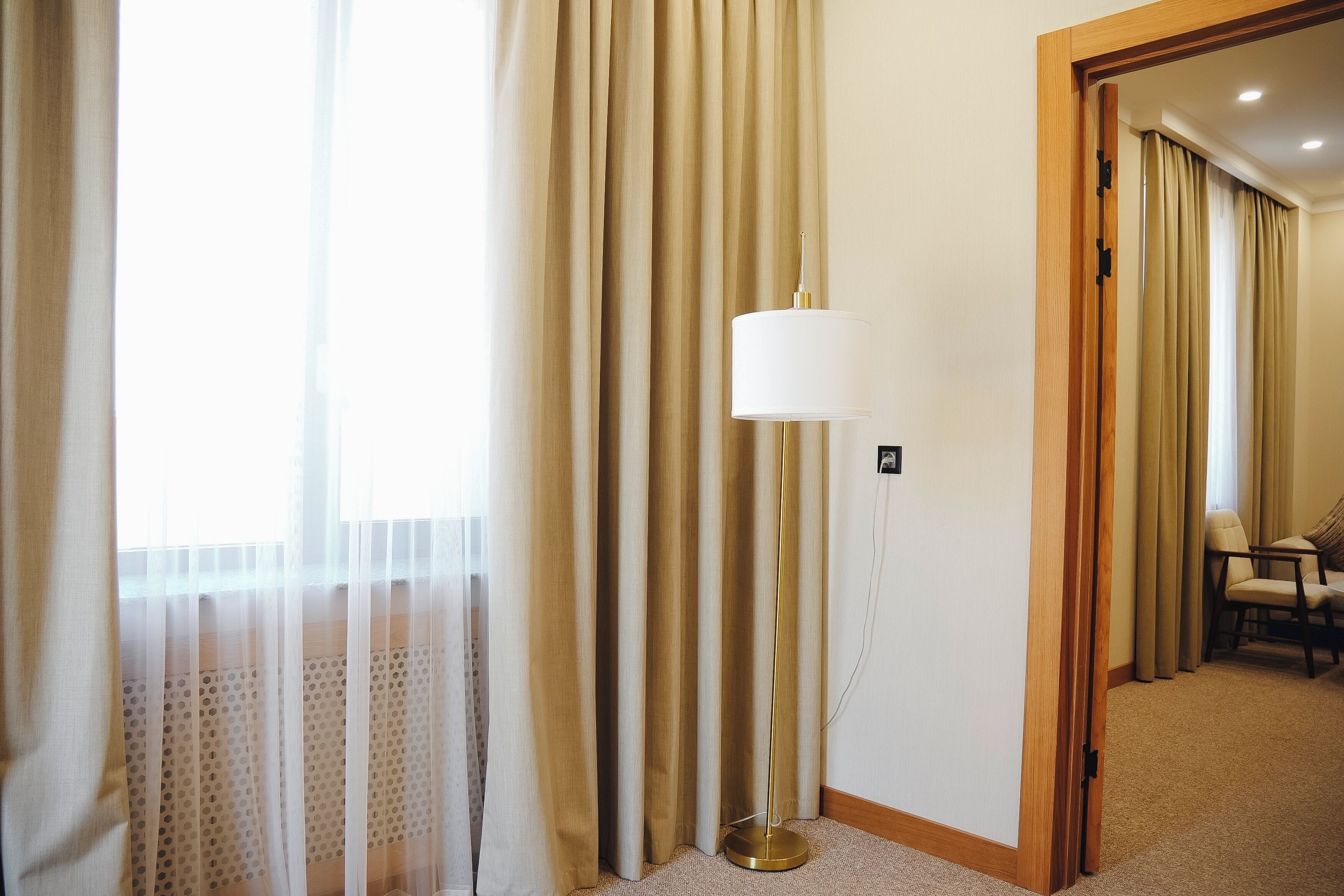 Quarto de hotel com abajur, cortinas bege e piso coberto por carpete.