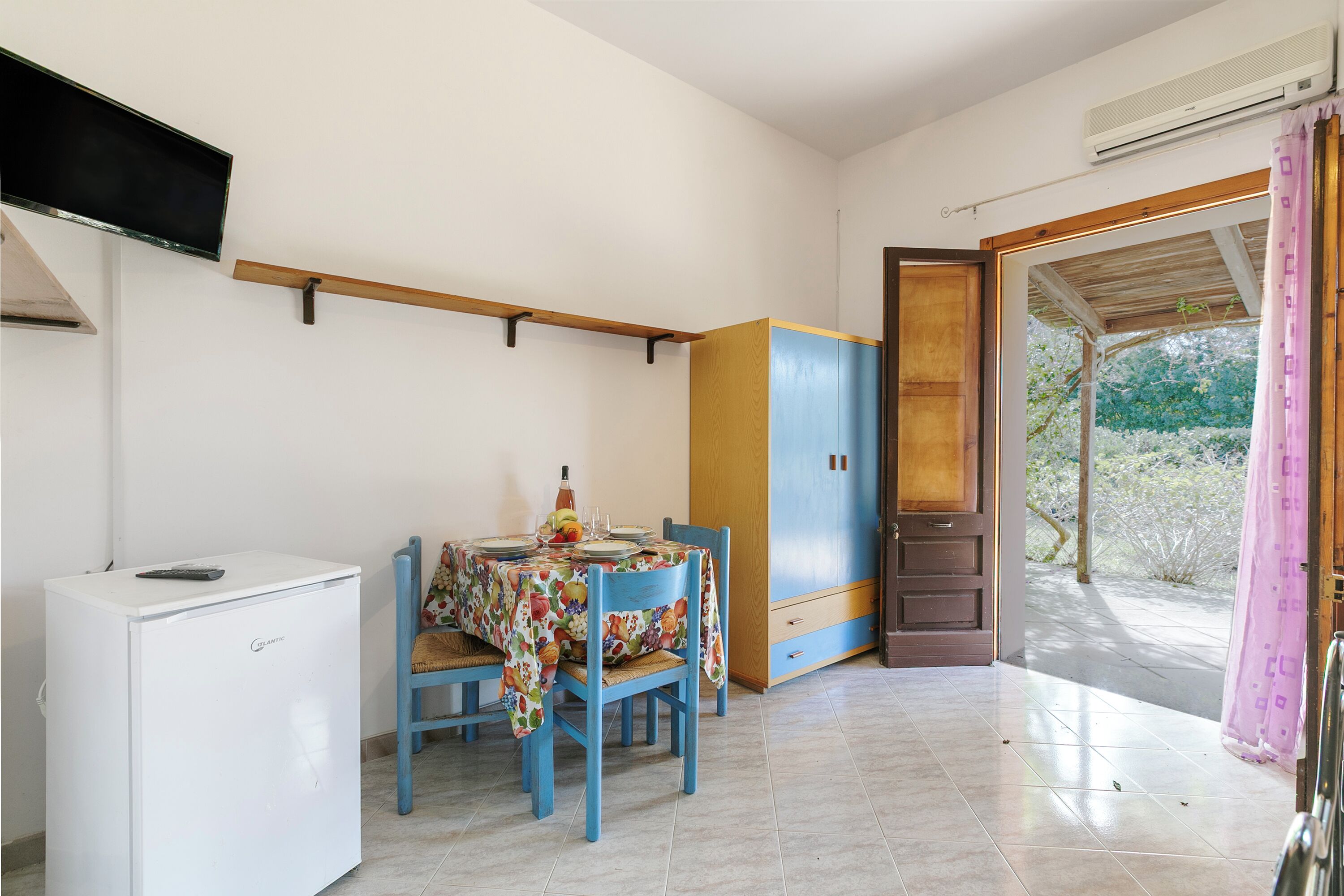 Cozinha compacta com área de refeições, geladeira branca, mesa posta com toalha de mesa florida e vista pela porta aberta para o exterior.