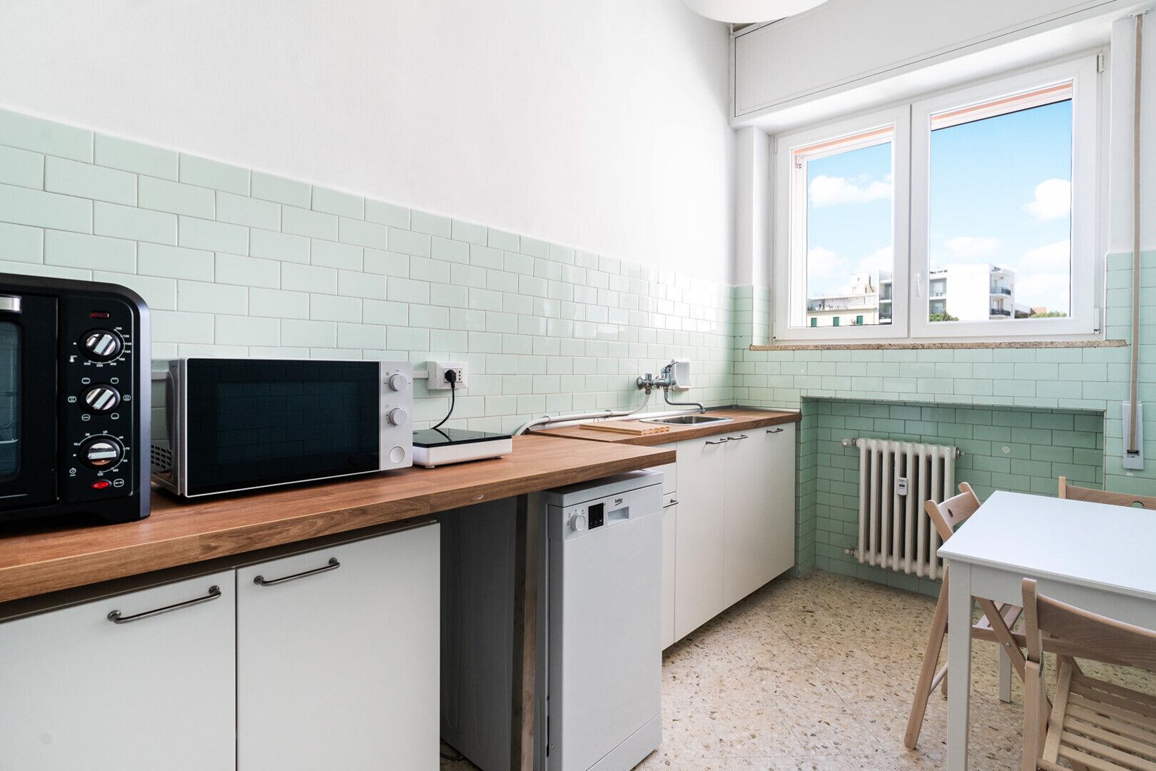 Cozinha compacta com armários brancos, bancada de madeira, micro-ondas, torradeira e janela com vista para edifícios.