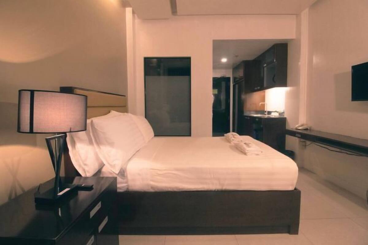 Quarto de hotel com cama, mesa de cabeceira, luminária e vista para a cozinha.