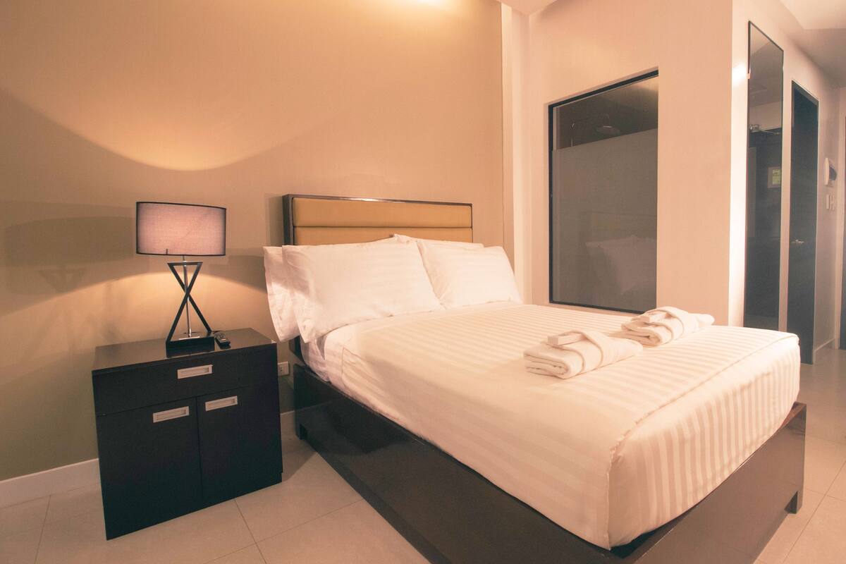 Cama bem arrumada com lençóis brancos e toalhas dobradas, mesa de cabeceira com abajur e cômoda escura.
