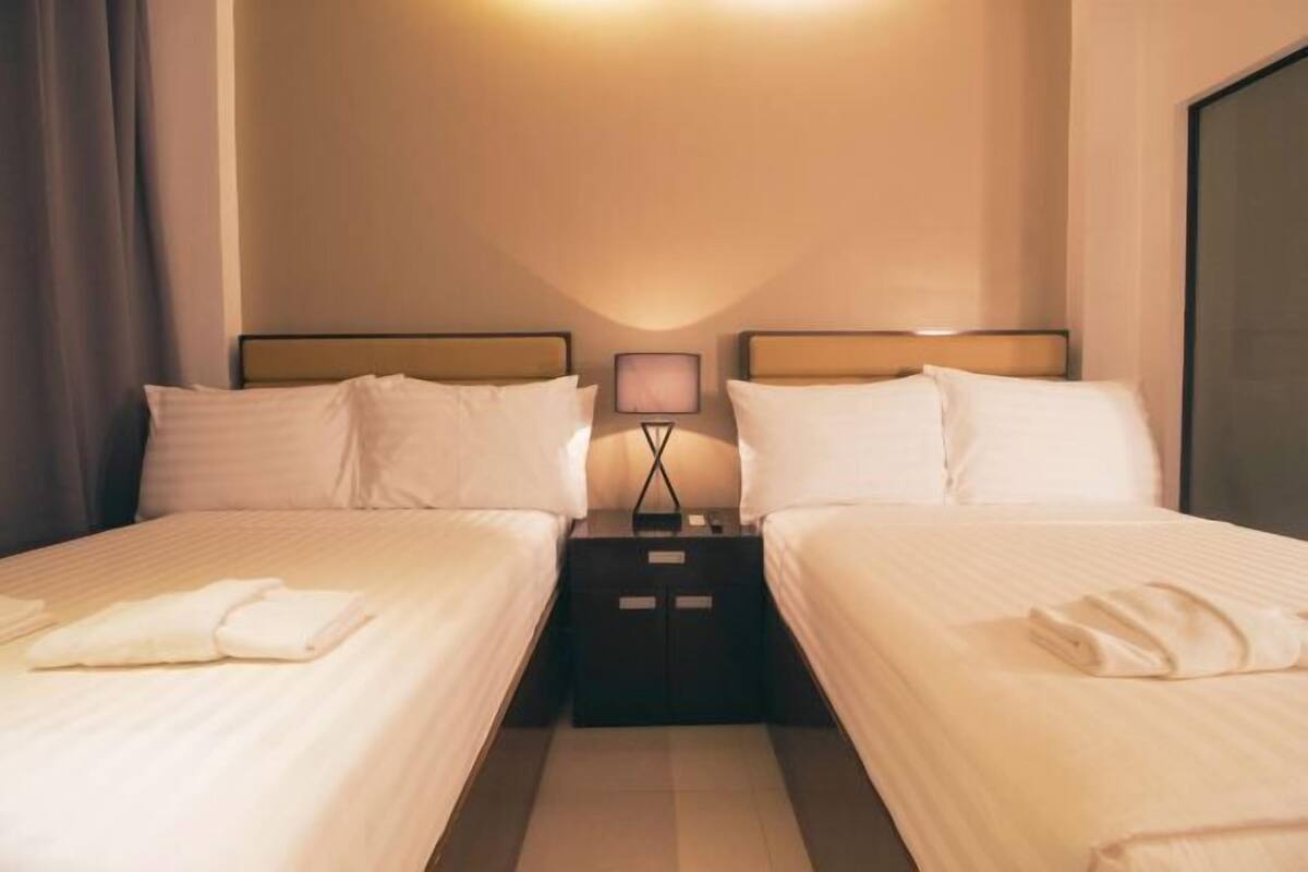 Quarto de hotel com duas camas, uma mesa de cabeceira com abajur e uma janela grande.