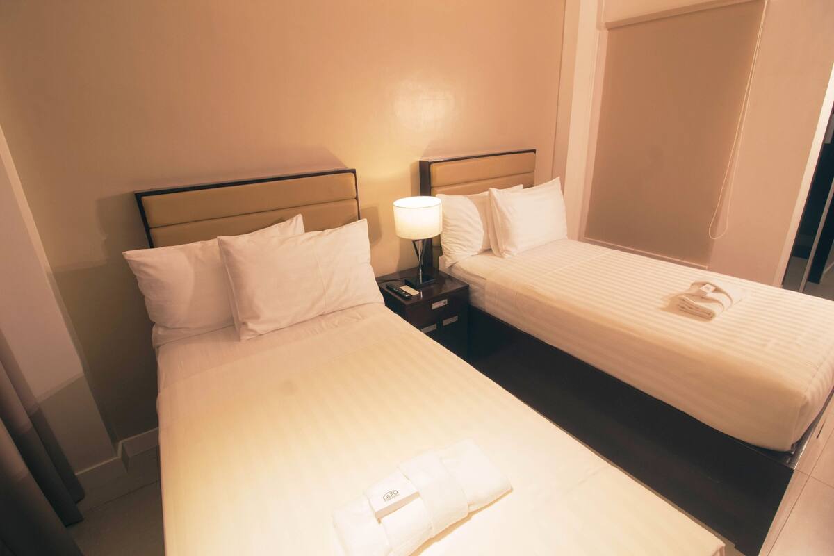 Quarto de hotel com duas camas, uma mesa de cabeceira com abajur e uma janela com persianas.