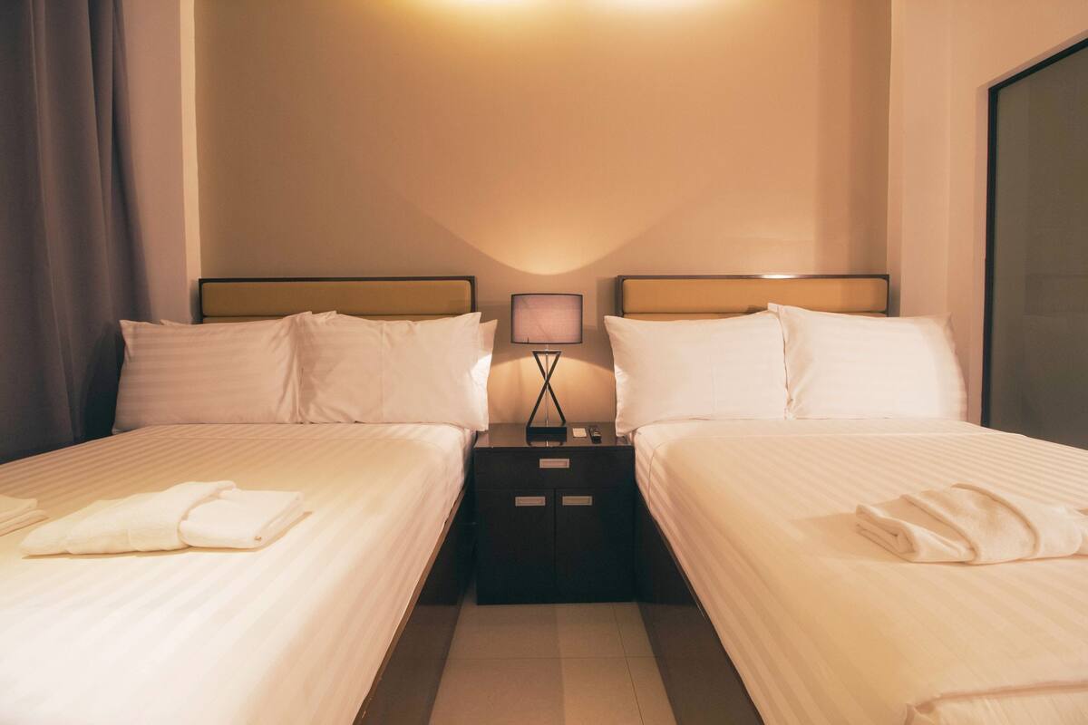 Quarto de hotel com duas camas, uma mesa de cabeceira com abajur e uma janela grande.