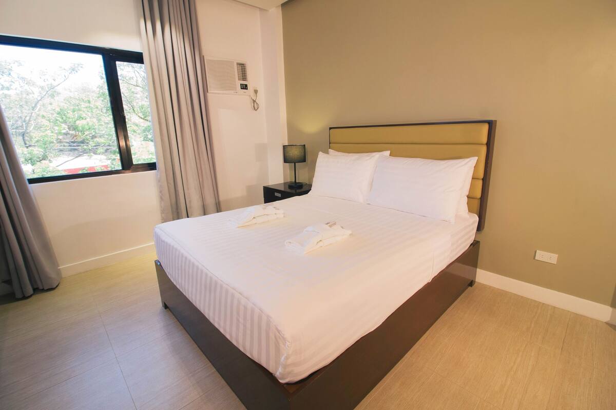 Cama bem arrumada com lençóis e toalhas brancas, mesa de cabeceira com abajur e janela com cortinas.