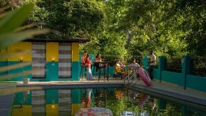 Uma área de piscina com um boião flutuante, uma estrutura pequena de paredes amarelas e verdes, e pessoas ao redor de uma mesa.