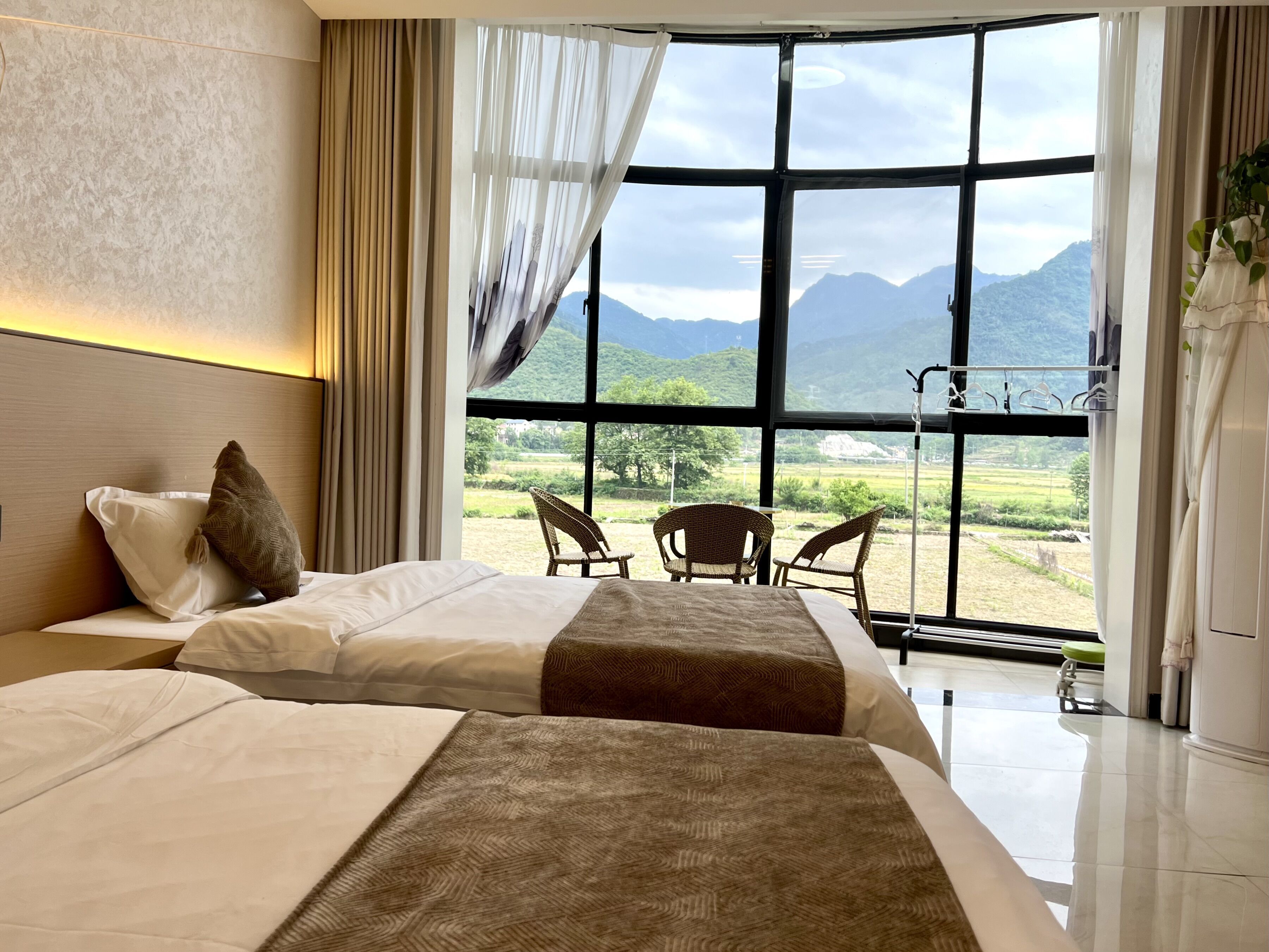 Quarto de hotel com cama, duas cadeiras, mesa e uma grande janela com vista para as montanhas.