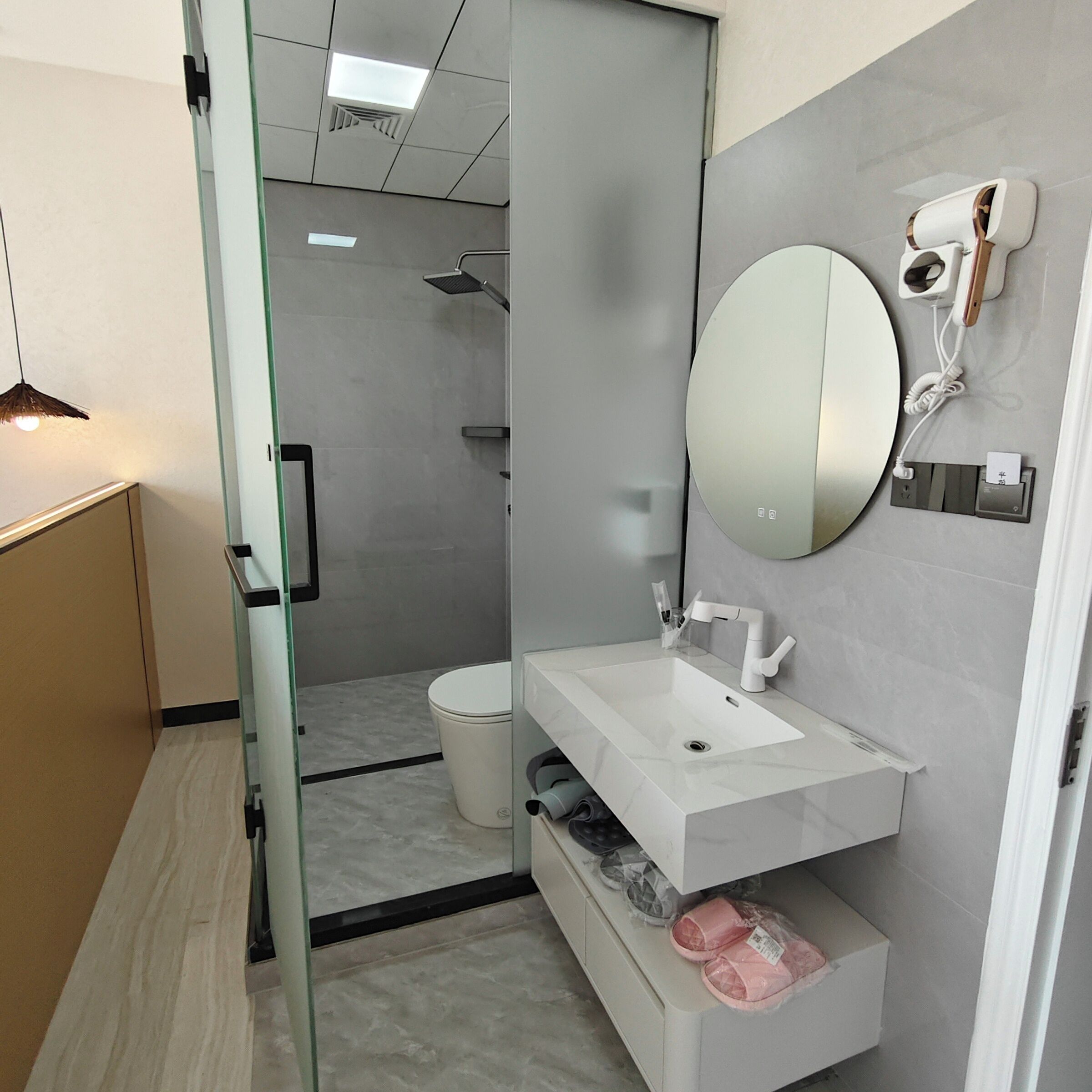 Uma casa de banho moderna com cabine de chuveiro em vidro, lavatório branco com espelho e secador de cabelo fixado na parede.