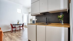Uma cozinha moderna com armários brancos, bancada de madeira e torneira preta. Há uma área de jantar com cadeiras vermelhas e uma mesa pequena.
