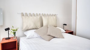 Uma cama bem arrumada com almofadas brancas e bege, uma mesinha de cabeceira com abajur, e um pequeno vaso com flores.