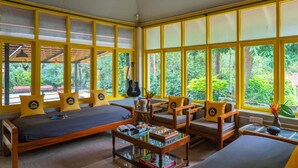 Quarto com cama, dois sofás, mesa de centro e janelas amplas com molduras amarelas.