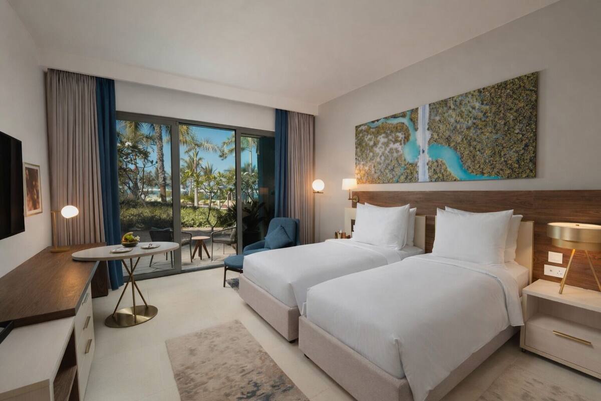 Quarto de hotel com duas camas, uma escrivaninha, uma cadeira e vista para palmeiras através de amplas janelas.