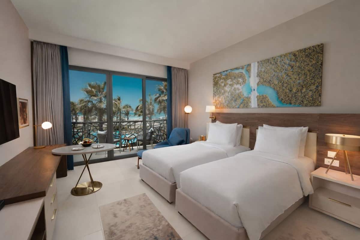 Quarto de hotel com duas camas, varanda com vista para a piscina, mesa de jantar e um quadro na parede.