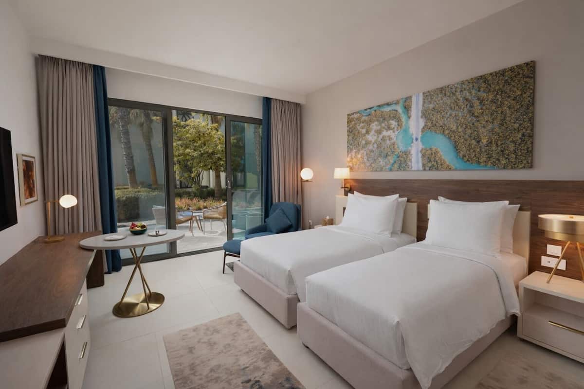 Quarto de hotel com duas camas, uma mesa de jantar, uma cadeira e uma janela grande com cortinas.