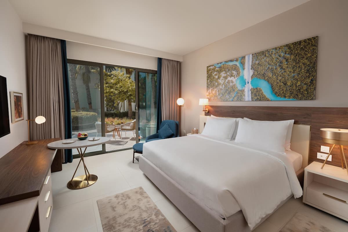 Quarto de hotel moderno com uma cama grande, uma escrivaninha, uma cadeira e vista para o exterior através de grandes janelas.