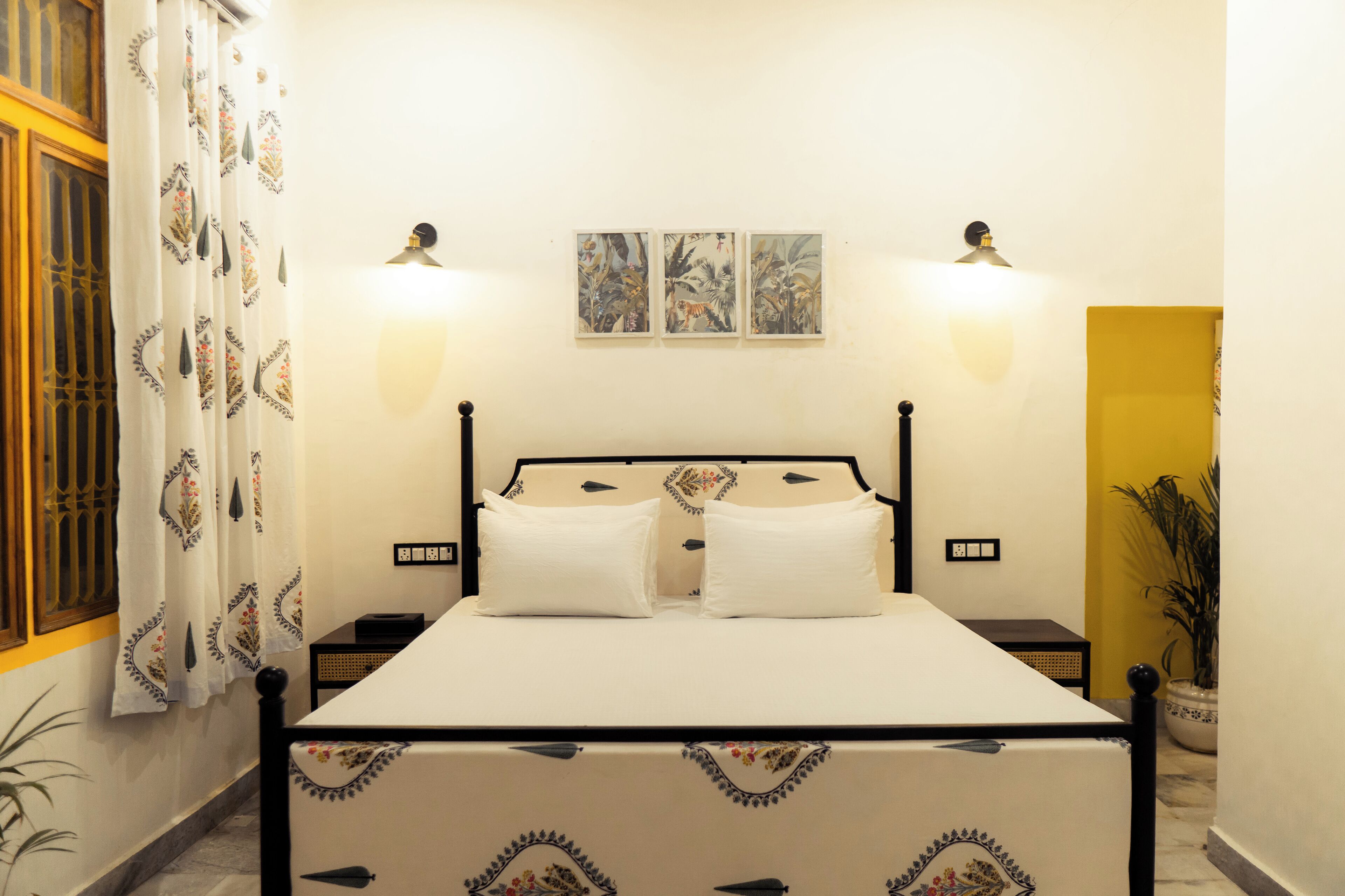 Cama bem arrumada com lençóis brancos e estrutura de metal preto, duas luminárias de leitura fixadas na parede e uma mesinha de cabeceira ao lado da cama.