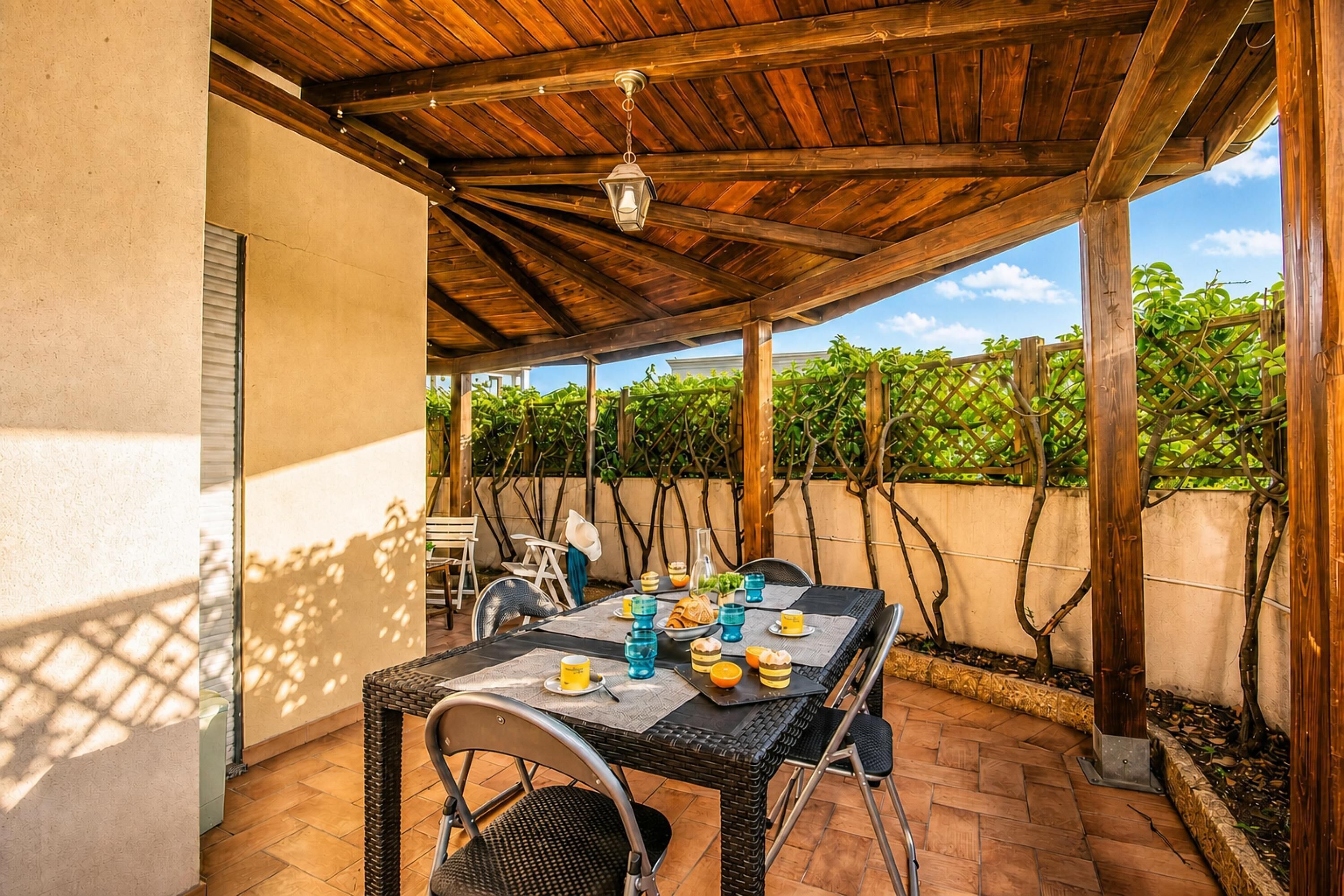Um pátio externo com mesa posta para refeições, rodeado por cadeiras e uma pérgula de madeira.