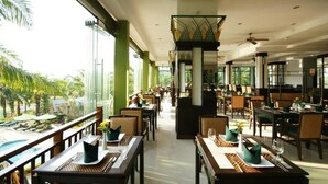 Um restaurante espaçoso com pé-direito alto, amplas janelas e vista para palmeiras. As mesas estão dispostas com guardanapos verdes e talheres.