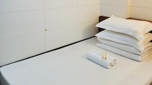 Uma cama bem arrumada com lençóis brancos e uma toalha enrolada, com uma faixa preta na lateral da cama, e uma parede de azulejos brancos.