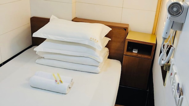 Uma cama bem arrumada com lençóis brancos, cabeceira de madeira, mesa de cabeceira com controle remoto e secador de cabelo na parede.