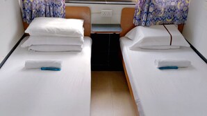 Duas camas de solteiro com lençóis brancos e toalhas azuis, uma mesinha e uma tomada elétrica entre elas.