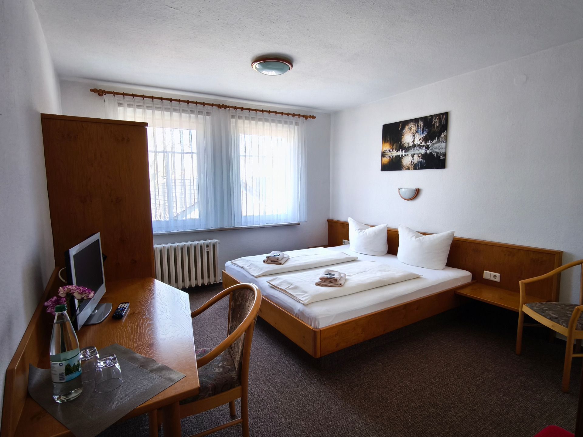 Quarto de hotel com cama de madeira, escrivaninha com computador e uma cadeira, janela com cortinas e um quadro na parede.