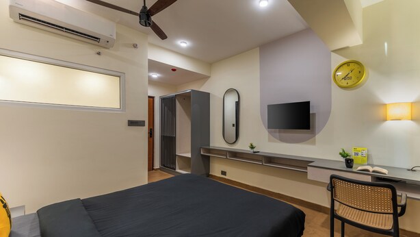 Quarto de hotel moderno com uma cama, uma escrivaninha com cadeira, uma televisão fixada na parede, um relógio e um aparelho de ar condicionado.