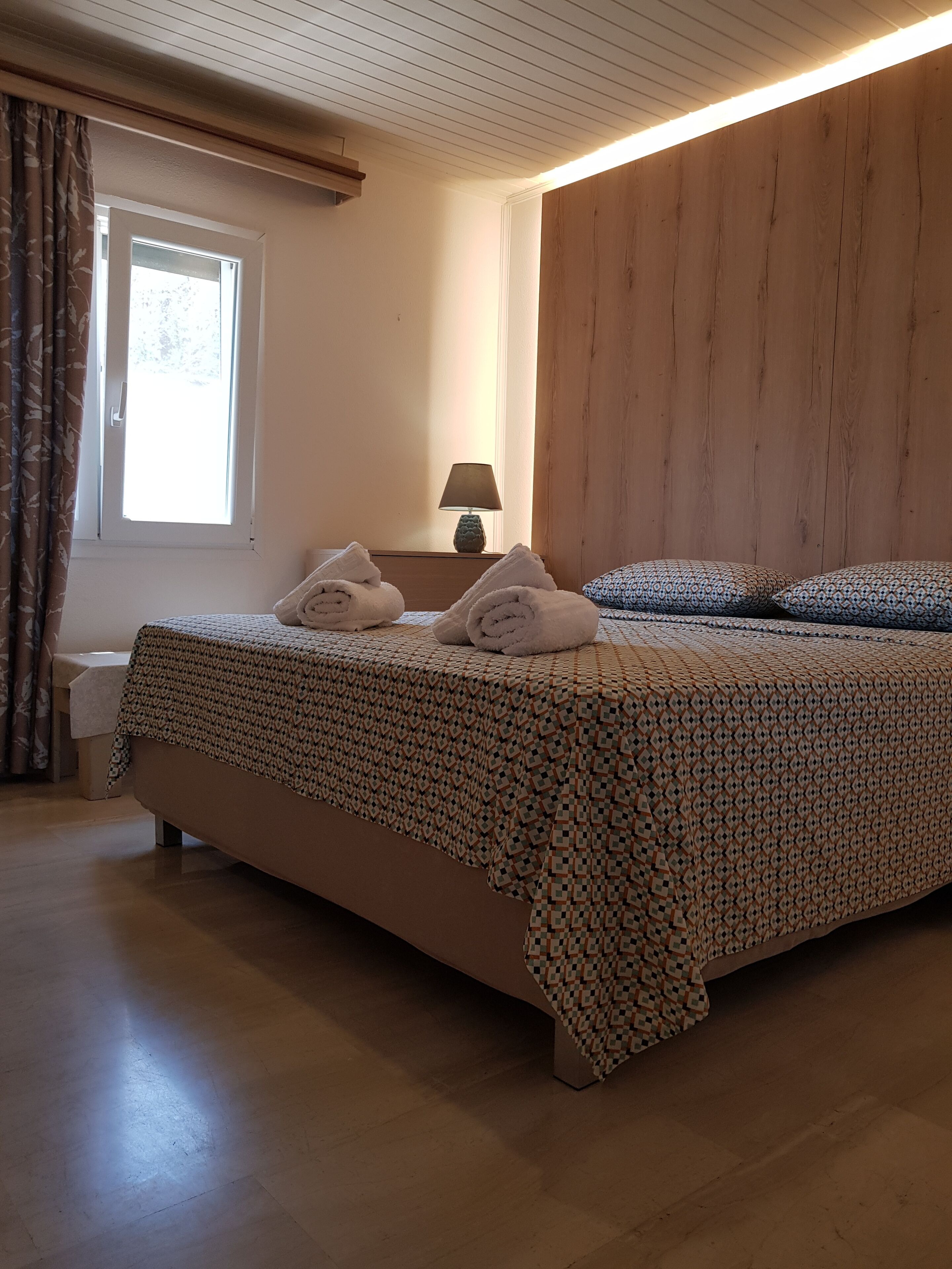 Uma cama bem arrumada com um cobreleite estampado, um abajur e uma janela com cortinas.