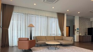 Uma sala de estar moderna com sofá, poltrona e mesa de centro, janelas amplas com cortinas e uma área de cozinha ao fundo.