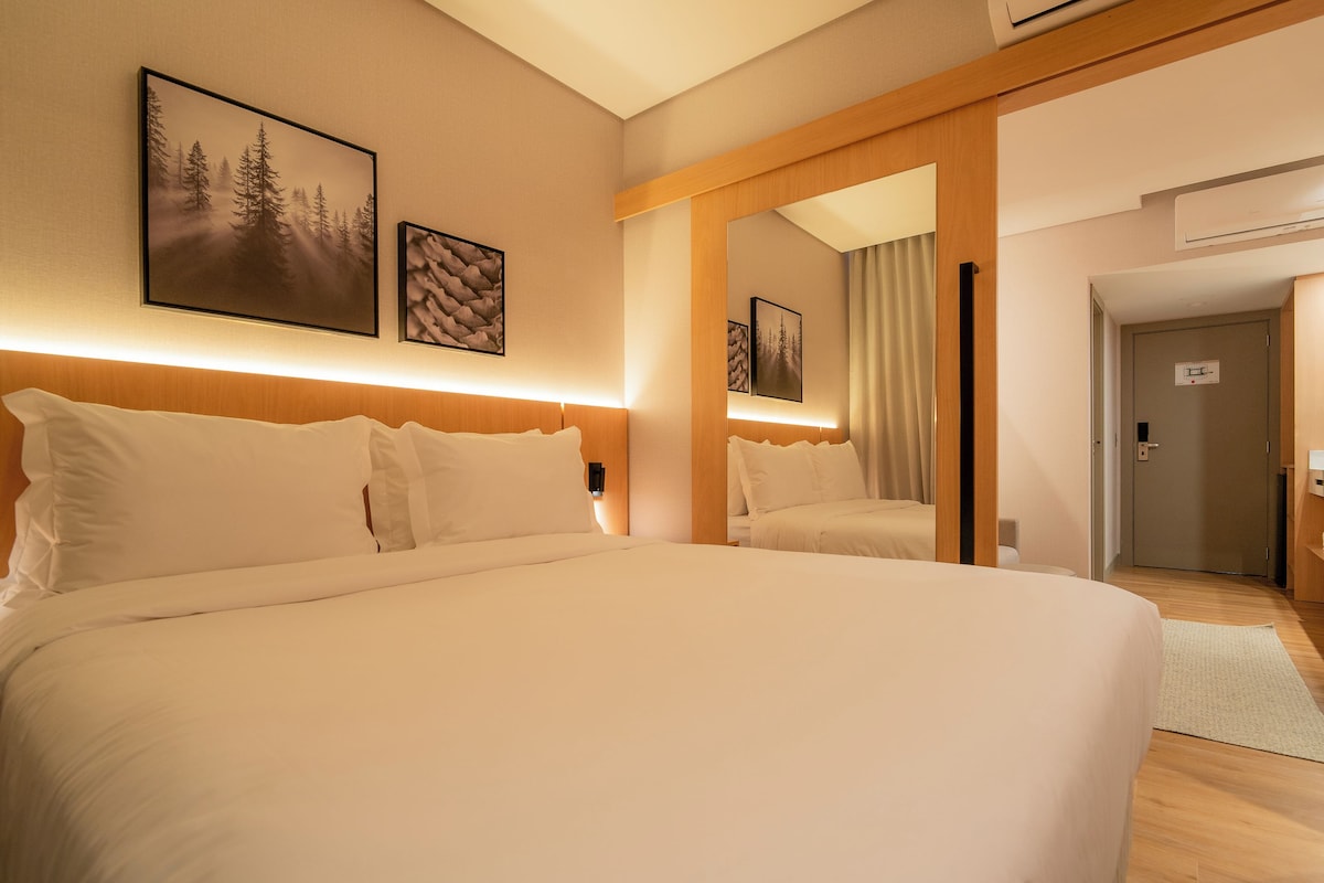 Quarto de hotel com uma cama grande, cabeceira de madeira e duas fotos emolduradas na parede.