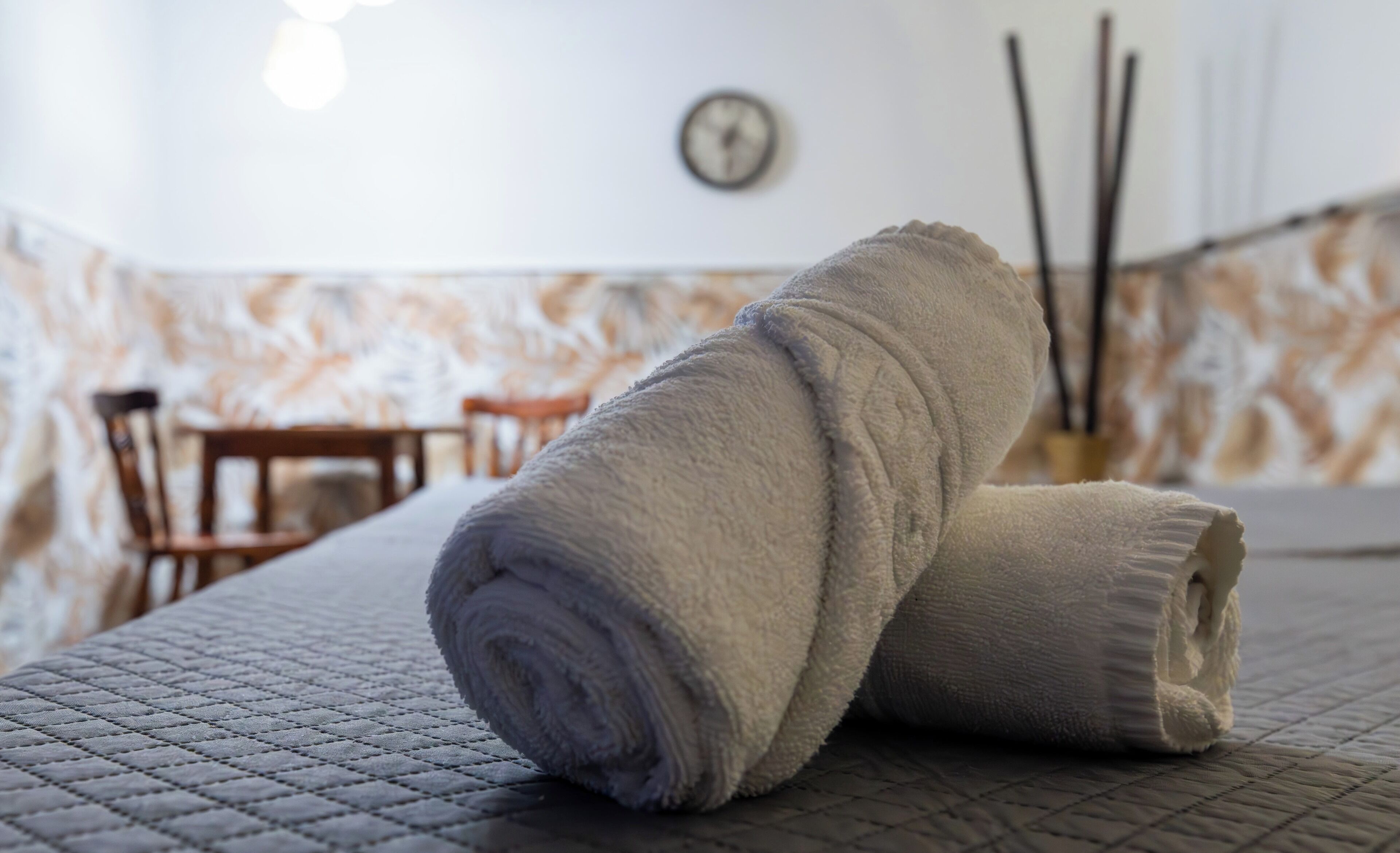 Uma toalha branca dobrada sobre um cobreleiro texturizado em um quarto com parede estampada, uma cadeira de madeira e um relógio na parede.