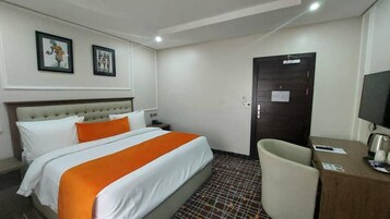 Quarto de hotel com cama, escrivaninha, cadeira, televisão e porta.