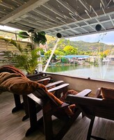 Restaurante com área externa e mesas ao ar livre, com vista para a água, apresentando mesas e cadeiras de madeira com almofadas.