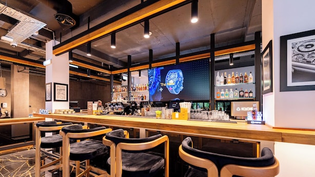 Uma área de bar com banquetas de madeira, um balcão com uma tela digital exibindo um relógio e prateleiras com diversos frascos.