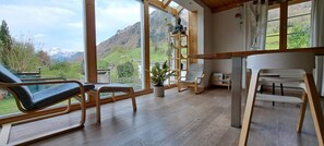 Quarto com janelas amplas, piso de madeira, sofá, poltrona, mesa pequena e estante com plantas.