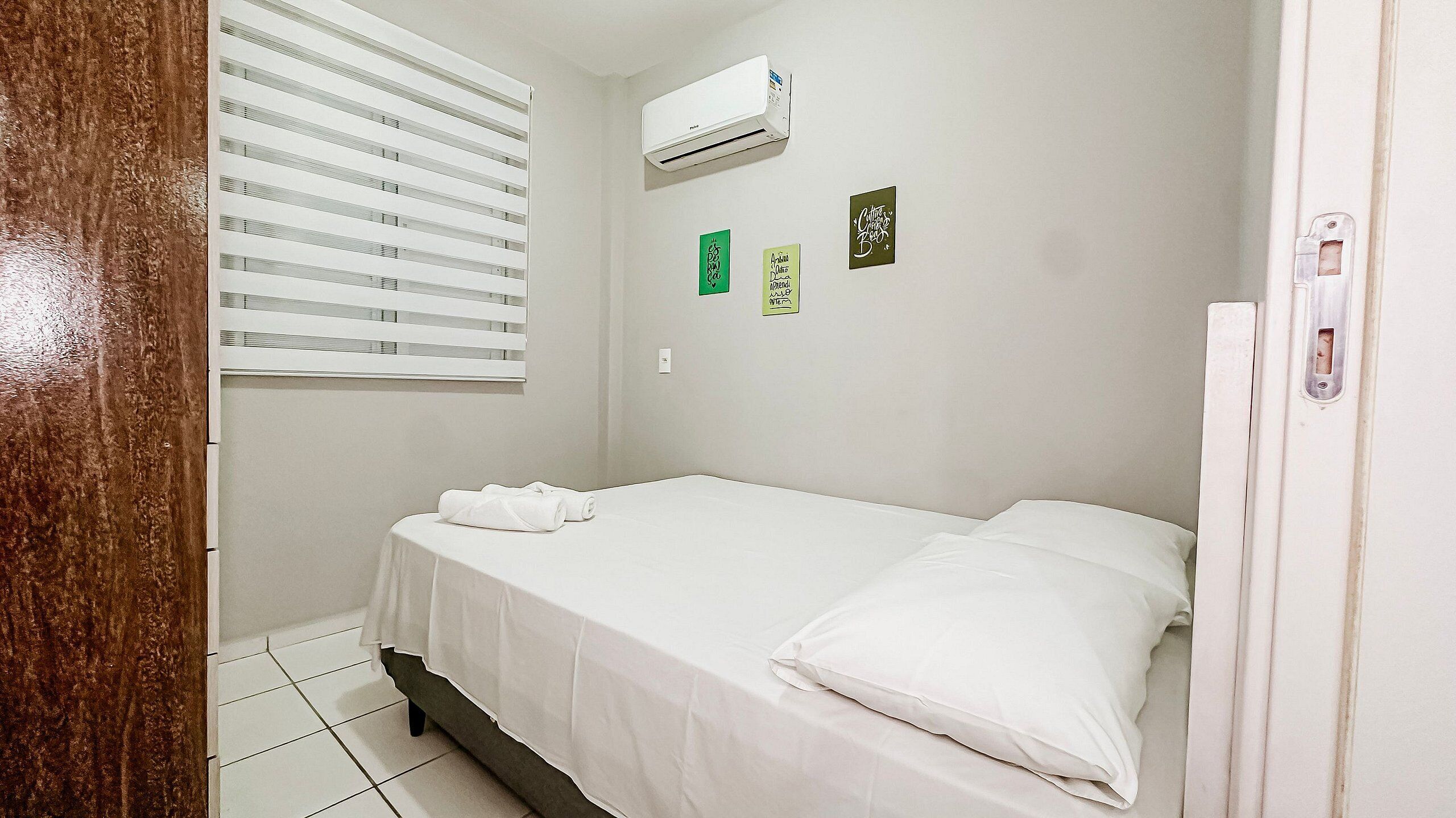 Quarto pequeno e limpo com uma cama de solteiro, janela com persianas, ar condicionado e algumas pequenas decorações nas paredes.