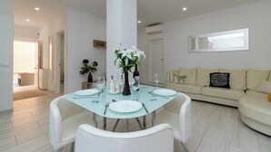 Uma sala de jantar moderna com mesa de vidro, cadeiras brancas e um pilar central. Há um sofá e um espelho na parede.