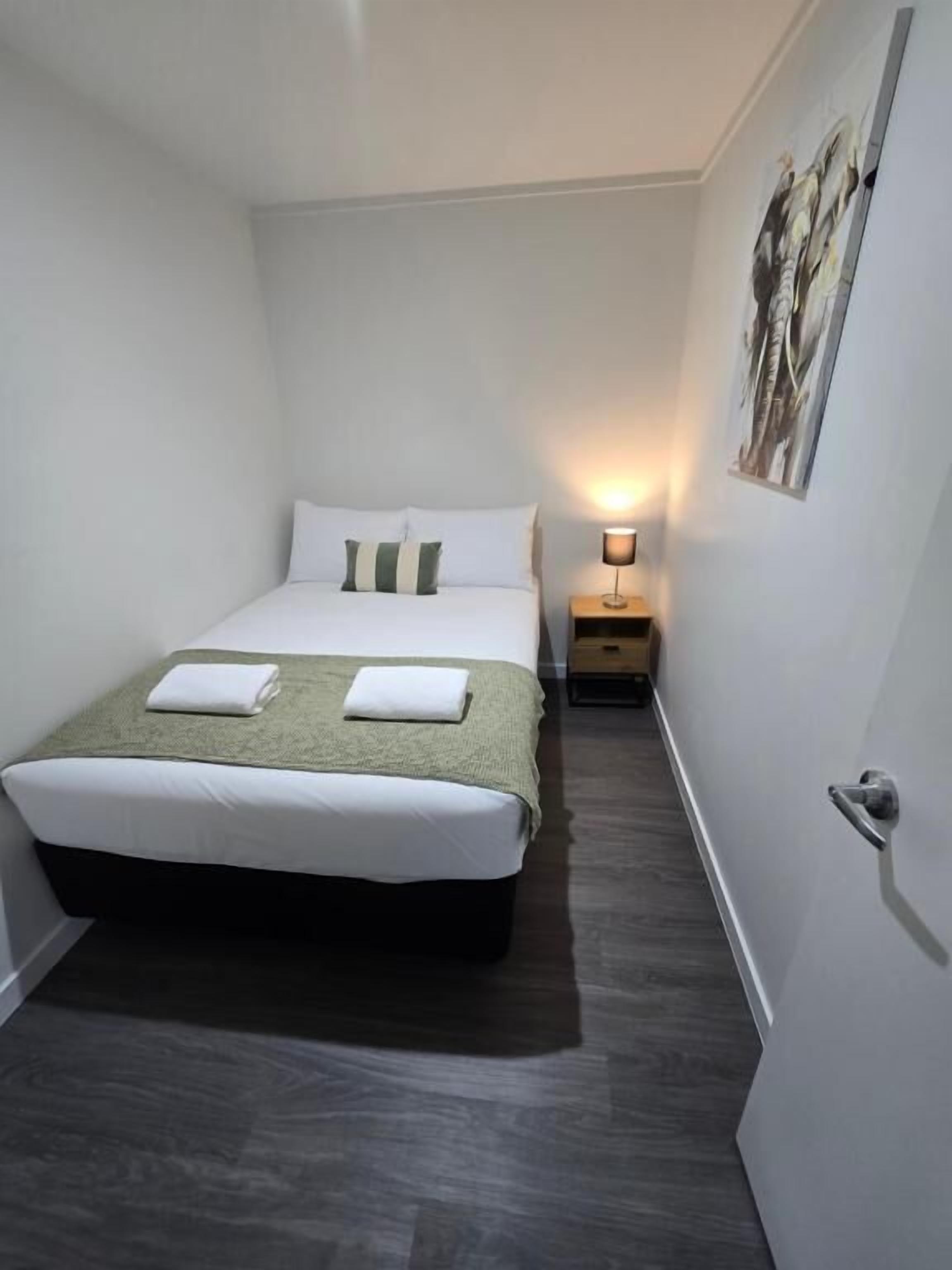 Um quarto pequeno e bem arrumado, com cama, travesseiros, mesa de cabeceira e abajur, e uma porta.