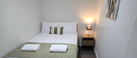 Um quarto pequeno e bem arrumado, com cama, travesseiros, mesa de cabeceira e abajur, e uma porta.