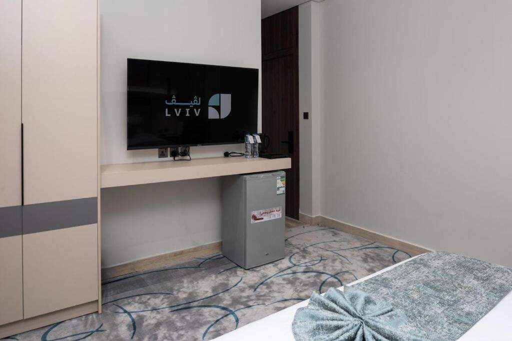 Quarto de hotel com televisão na parede, um pequeno frigorífico, uma escrivaninha com cadeira e uma cama com manta.